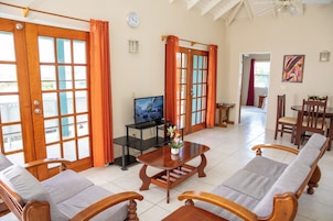 Uma sala de estar com televisão, mobiliário de madeira e grandes janelas com cortinas.