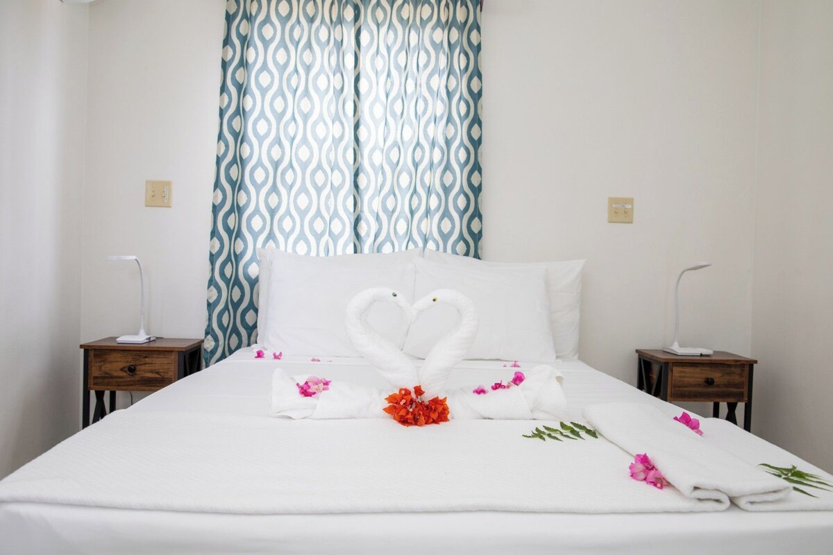 Quarto de hotel com uma cama, duas mesas de cabeceira, uma toalha decorativa em formato de cisne e cortina estampada.