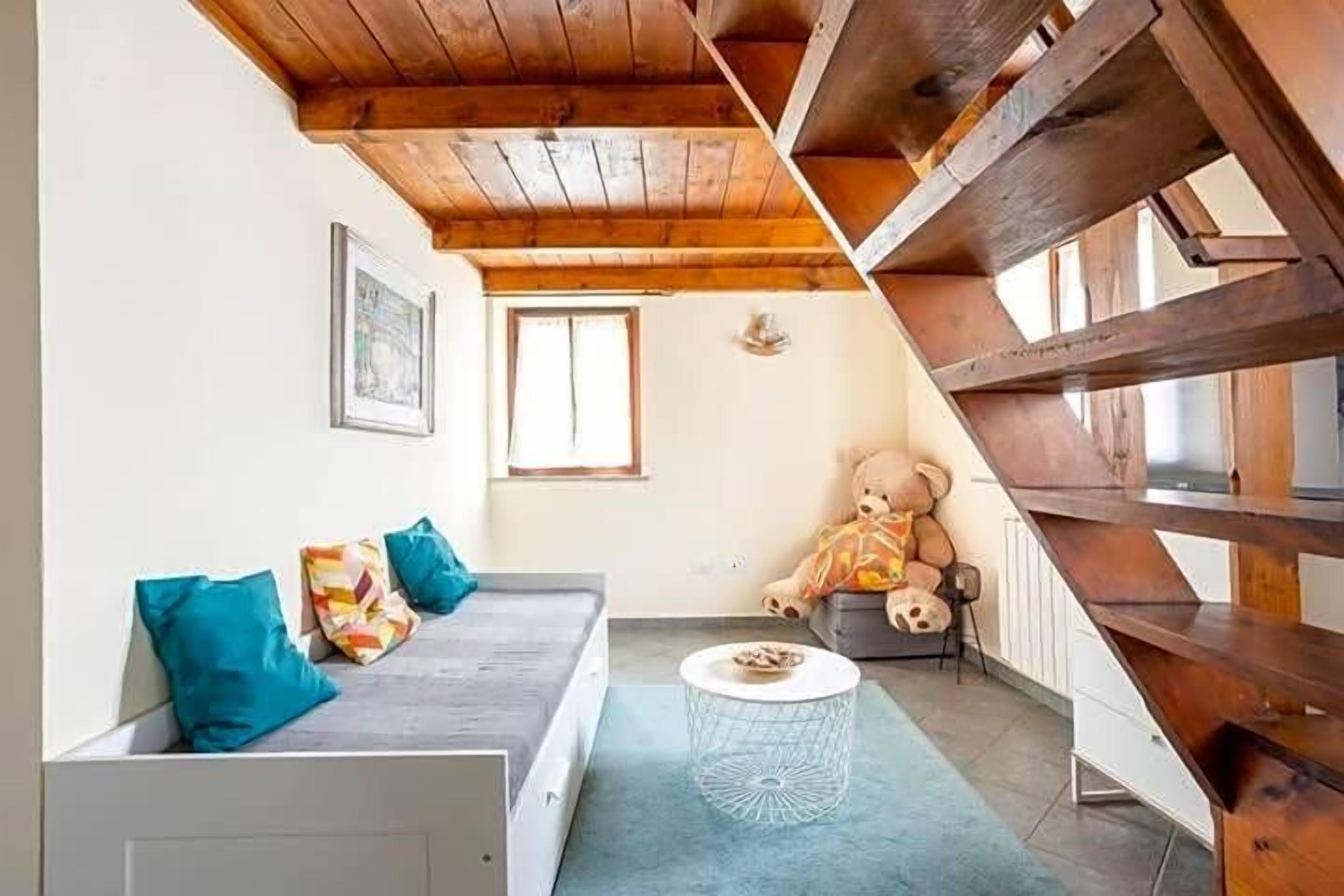 Quarto com teto de madeira, sofá com almofadas, mesa redonda pequena e um urso de pelúcia.
