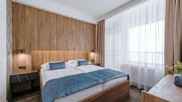 Quarto de hotel moderno com cabeceira de madeira, cama grande com manta azul, duas mesas de cabeceira com abajures e vista para o exterior através de cortinas translúcidas.