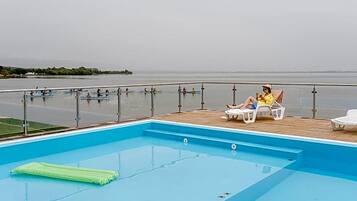 Piscina com vista para o mar, área de estar com cadeiras e uma pessoa relaxando.