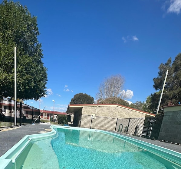 Uma piscina com borda curva, cercada por uma grade, e um prédio com telhado vermelho ao fundo.