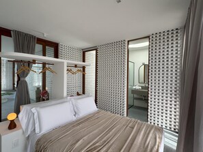 Quarto de hotel com cama, mesa de cabeceira com abajur, guarda-roupa, espelho e banheiro visível através de uma porta aberta.