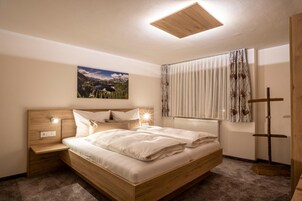 Quarto com cama de madeira, mesa de cabeceira, luminária na parede, janela com cortinas leves e uma pintura de paisagem.