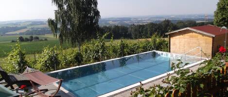 Piscina azul e límpida com vista para o campo, rodeada por vegetação e uma estrutura de madeira.