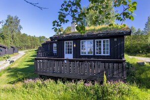 Uma cabana pequena com deck de madeira e uma porta, rodeada por vegetação e um céu limpo.