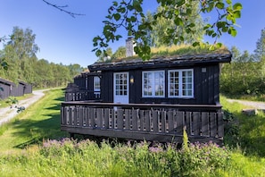 Uma cabana pequena com deck de madeira e uma porta, rodeada por vegetação e um céu limpo.