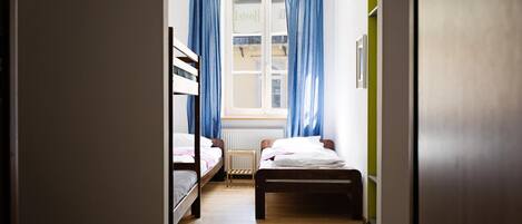 Quarto em dormitório com beliches, janela com cortinas, radiador e uma pequena mesa.