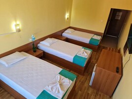 Quarto de hotel com três camas de solteiro, uma escrivaninha de madeira, uma mesinha pequena e uma televisão presa à parede.