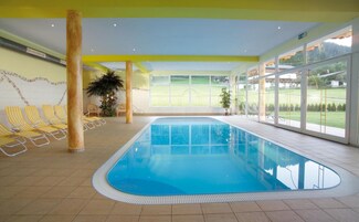 Piscina interna com paredes de vidro transparente, cadeiras de sol listradas de amarelo e vista para uma paisagem verde.