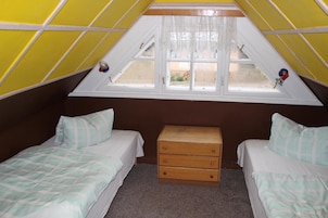 Um quarto de sótão pequeno e triangular com duas camas, uma cômoda de madeira e uma janela com vista para o exterior.