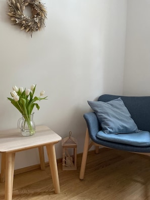 Um quarto com uma mesa de madeira, um vaso com flores brancas, um sofá azul e um pequeno banquinho de madeira.