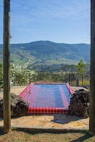 Piscina com deck ladrilhado em vermelho e azul, rodeada por uma cerca de metal, com colinas suaves e céu claro ao fundo.