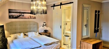 Quarto com cama grande, mesa de cabeceira, luminária na parede, quadro e porta deslizante para um banheiro.