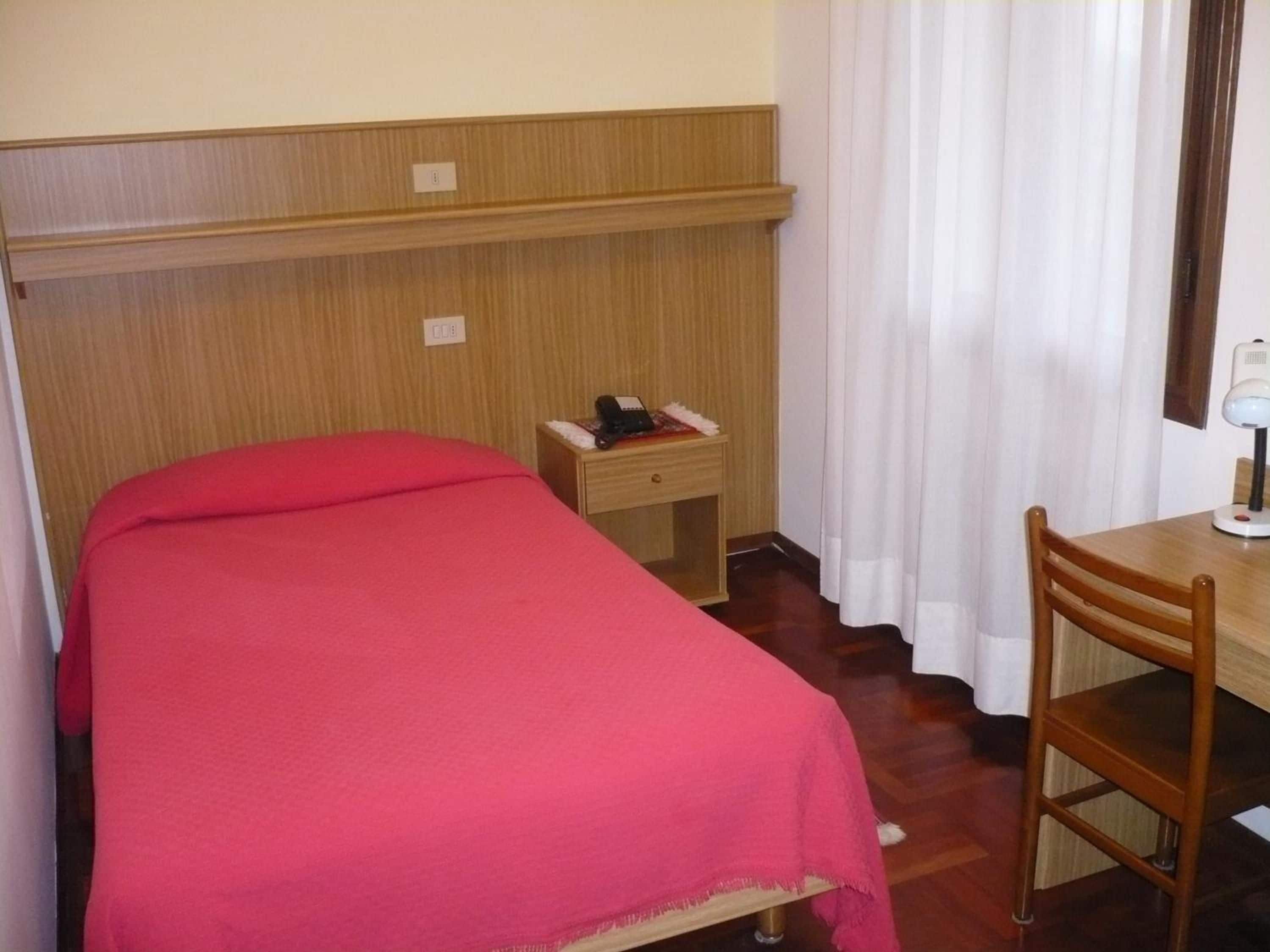 Quarto de hotel com cama, escrivaninha, cadeira, mesa de cabeceira e abajur.