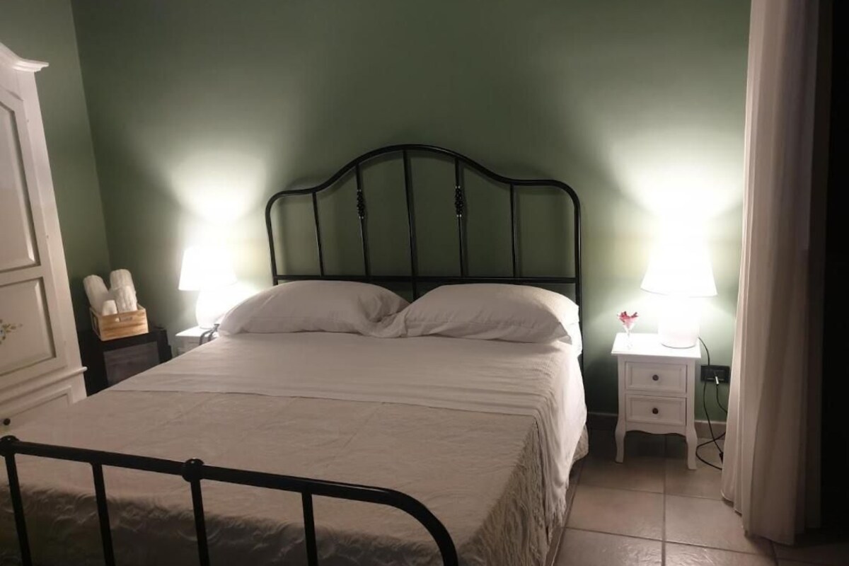Uma cama com estrutura de metal, uma mesa de cabeceira com abajur e outro criado-mudo com luminária.