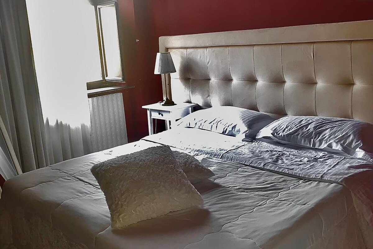 Uma cama com cabeceira branca e um travesseiro, uma mesa de cabeceira com luminária e uma janela com cortinas.