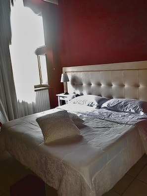 Uma cama com cabeceira branca e um travesseiro, uma mesa de cabeceira com luminária e uma janela com cortinas.