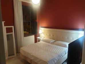 Quarto com cama, mesa de cabeceira, abajur e janela com cortinas.
