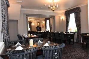Uma sala de jantar de hotel com mesas preparadas para quatro pessoas, cadeiras estofadas pretas, um candelabro e cortinas estampadas.
