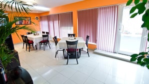 Uma área de refeições com mesa e cadeiras brancas, paredes laranja e janelas amplas com cortinas cor-de-rosa.