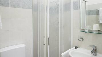 Casa de banho com chuveiro em cabine de vidro, lavatório branco e prateleira de vidro com produtos de higiene pessoal.