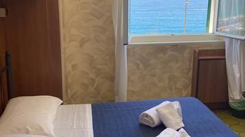 Uma cama com roupa de cama branca e azul, uma janela com vista para o mar e uma cortina com padrão de folhas.