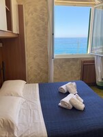 Uma cama com roupa de cama branca e azul, uma janela com vista para o mar e uma cortina com padrão de folhas.
