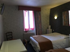 Quarto de hotel com duas camas, uma mesa, uma cadeira, um radiador e uma janela com cortinas.