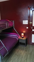 Uma cama beliche com roupa de cama roxa, uma pequena mesa de cabeceira com abajur e uma porta de madeira.