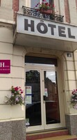 Entrada de hotel com um letreiro grande acima da porta, uma placa na parede e floreiras em ambos os lados.