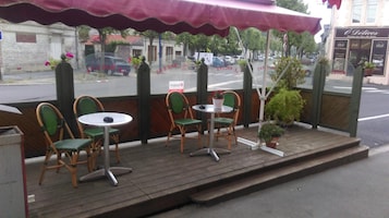 Área de estar externa com cadeiras e mesas de vime, toldo coberto e vista para a rua com carros estacionados e edifícios.