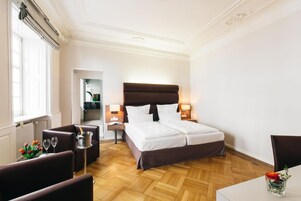 Quarto de hotel com cama de casal, mesas de cabeceira, área de estar com uma poltrona, uma mesinha e um vaso com flores.