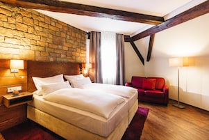 Quarto de hotel com parede de pedra, vigas de madeira, cama com roupa branca, sofá vermelho e luminária de chão.