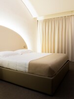 Quarto de hotel com uma cama de solteiro, cabeceira, janela com cortinas e piso coberto por carpete.