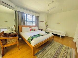 Quarto de hotel com cama de madeira, escrivaninha com cadeira, mesa de cabeceira, janela com cortinas e ventoinha no teto.
