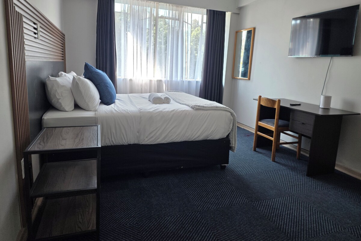 Quarto de hotel com cama, escrivaninha, cadeira, televisão de tela plana e janela com cortinas.