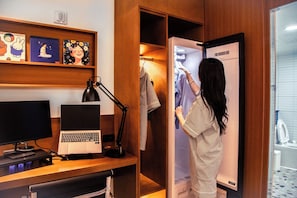 Quarto de hotel com escrivaninha, computador, luminária e guarda-roupa com roupas.