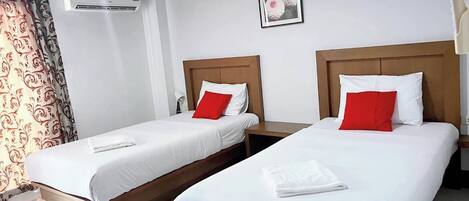 Quarto de hotel com duas camas de solteiro, cada uma com cabeceira de madeira, roupa de cama branca e almofada vermelha. Há um quadro na parede acima das camas, e um aparelho de ar condicionado fixado na parede.