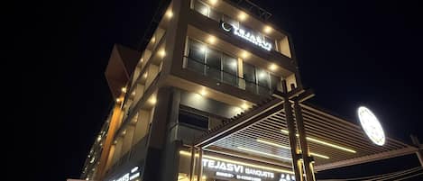 Um edifício com letreiros iluminados para 'TEJASVI BANQUETS' e 'SWAD FOOD LAND' à noite.