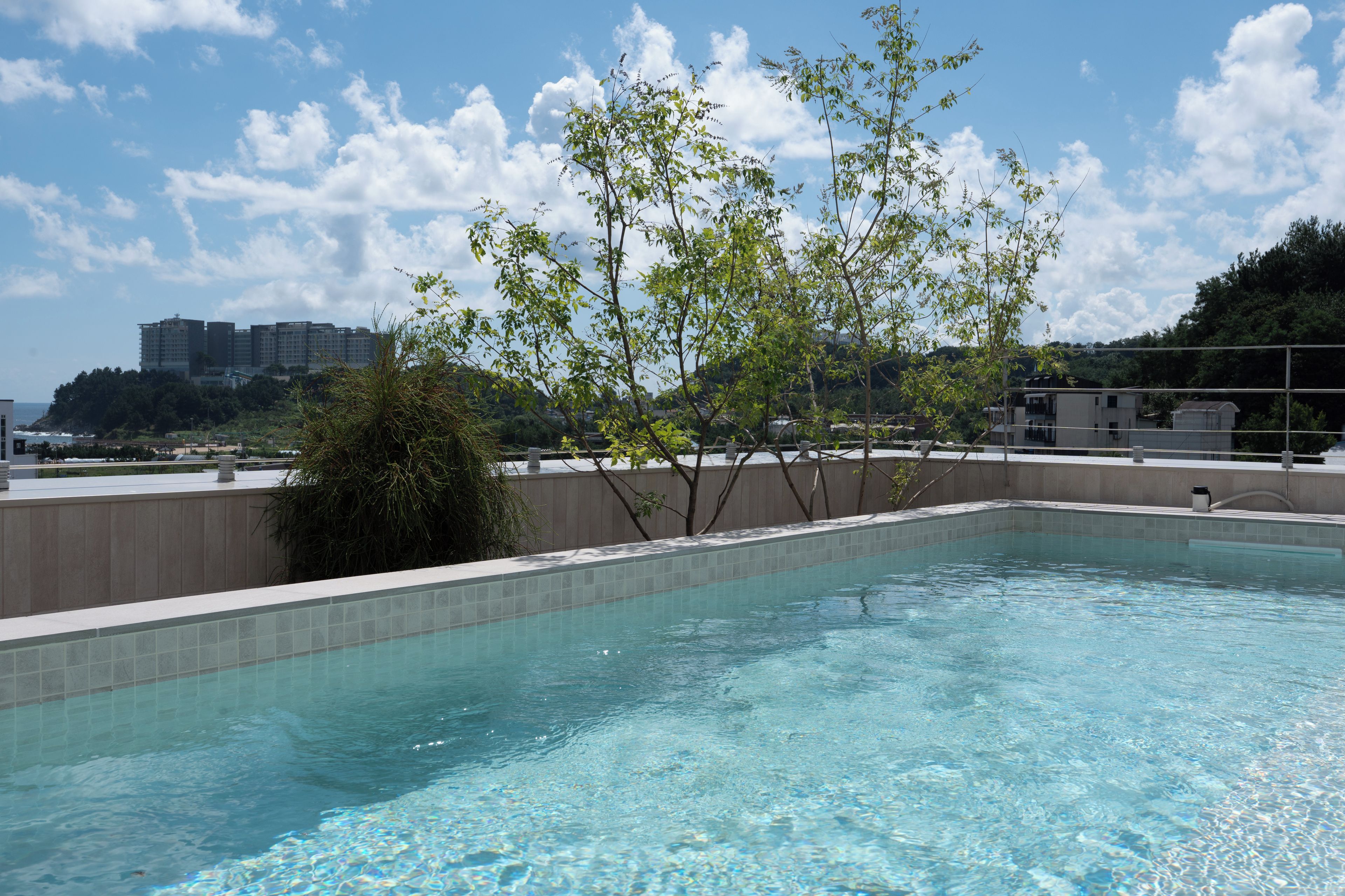 Piscina com água cristalina, rodeada por um deck revestido e um muro baixo. Ao fundo, edifícios e árvores.