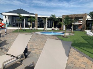 Área de piscina com espreguiçadeiras, uma piscina pequena e um edifício com janelas amplas e colunas de pedra.