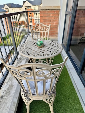 Uma varanda com mesa redonda e cadeiras, porta de vidro e vista para edifícios ao fundo.