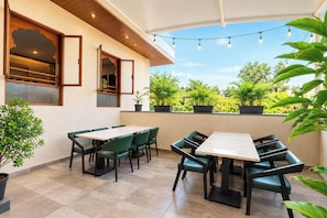 Um pátio externo com mesa e cadeiras, plantas em vasos e luzes de cordão.
