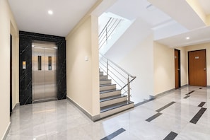 Interior moderno com elevador de mármore, escada com guarda-corpo de metal e corredor que leva a uma porta com a inscrição '1007'.