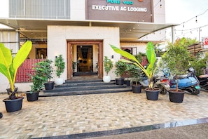 Entrada de hotel com calçada de azulejos, plantas em vasos e uma motocicleta estacionada na frente.