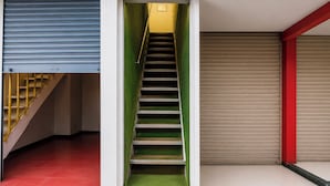Uma escadaria com carpete verde leva a um patamar, ladeada por persianas venezianas fechadas.