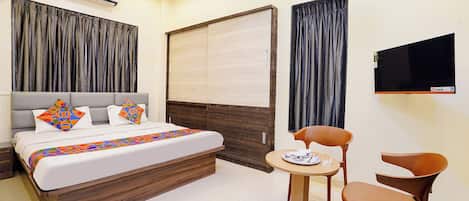 Quarto de hotel com cama, televisão, mesa pequena e cadeiras, e janela com cortinas.