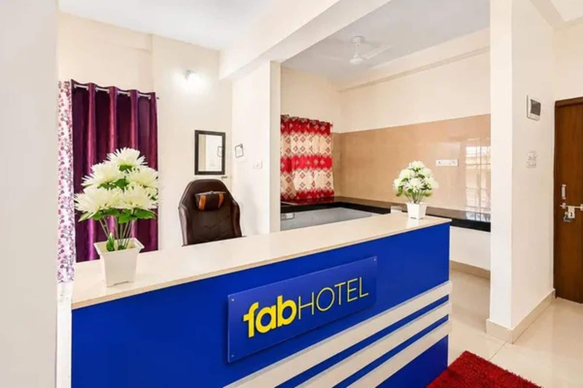 Balcão de recepção de hotel com placa azul, uma cadeira, uma mesa, cortina vermelha e um vaso com flores.
