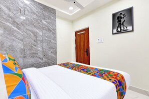 Quarto de hotel com uma cama, porta de madeira, luminária fixada na parede e um quadro emoldurado.
